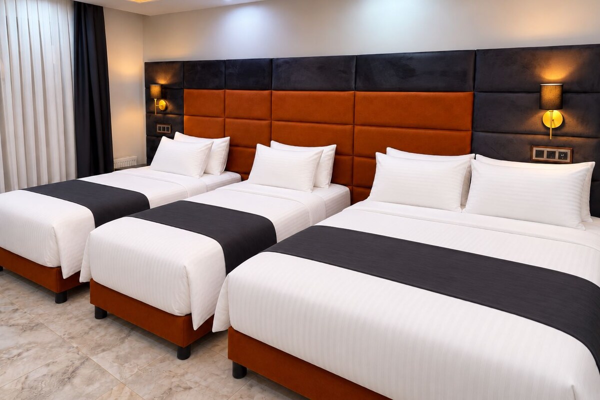 Quarto de hotel com três camas, uma cabeceira grande e luminárias fixadas na parede.