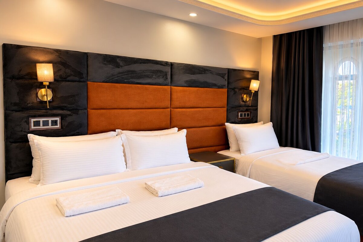 Quarto de hotel com duas camas, cabeceira moderna, abajures e vista para o verde pela janela.