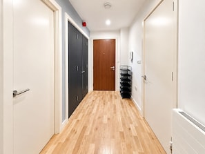Um corredor com piso de madeira, uma porta de madeira no final e uma estante de metal preto com prateleiras.