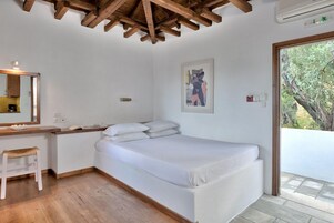 Quarto com teto de madeira, cama, um banquinho pequeno, espelho e janela com vista para árvores.