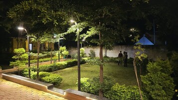 Área externa bem iluminada com um caminho pavimentado, arbustos bem aparados e um grupo de pessoas reunidas perto de um muro.