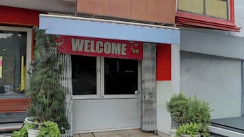 Fachada de loja com uma placa 'WELCOME' sobre a entrada, ladeada por plantas em vasos.