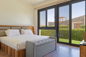 Quarto com cama grande, cabeceira de madeira e vista para uma parede de pedra e vegetação lá fora.