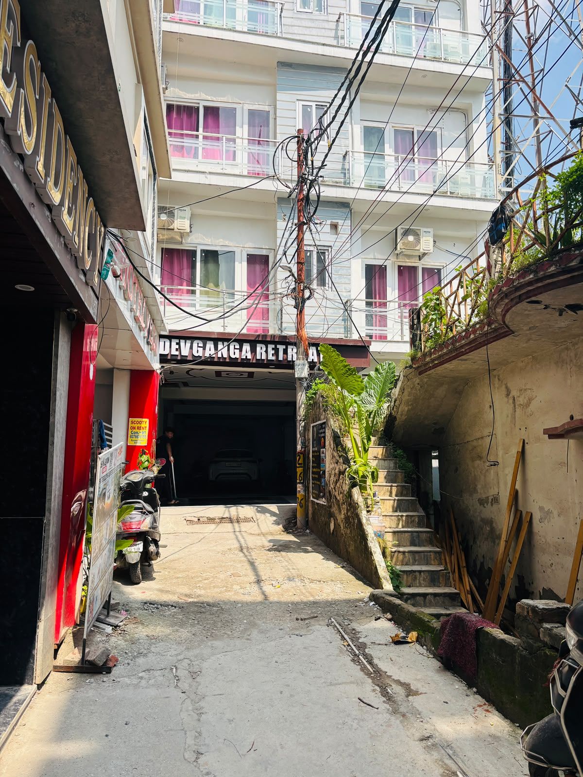 Uma rua estreita com um edifício identificado como 'DEVANGA RETREAT' e uma scooter estacionada.