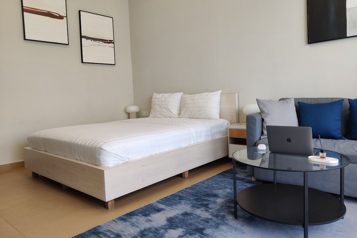 Quarto moderno com cama, mesas de cabeceira, um laptop sobre uma mesa de vidro e um sofá com almofadas.