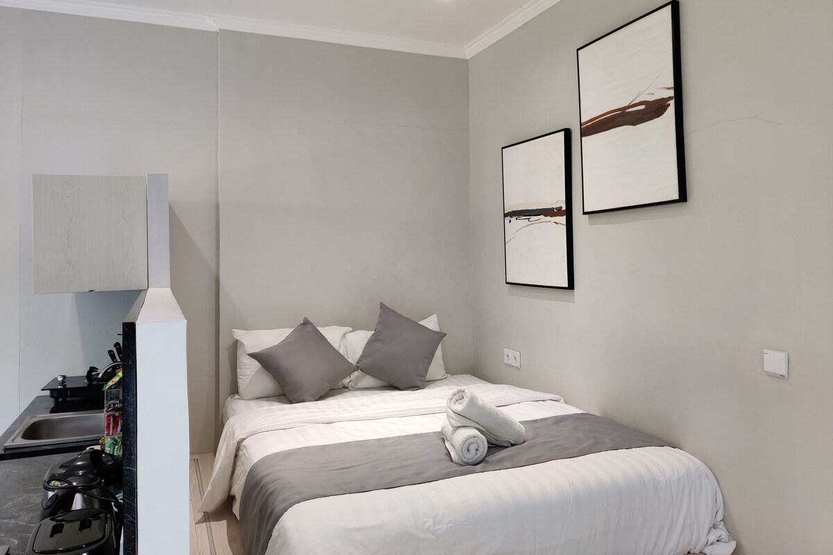 Quarto com cama, almofadas, toalha, obra de arte na parede e um espaço de cozinha compacta.