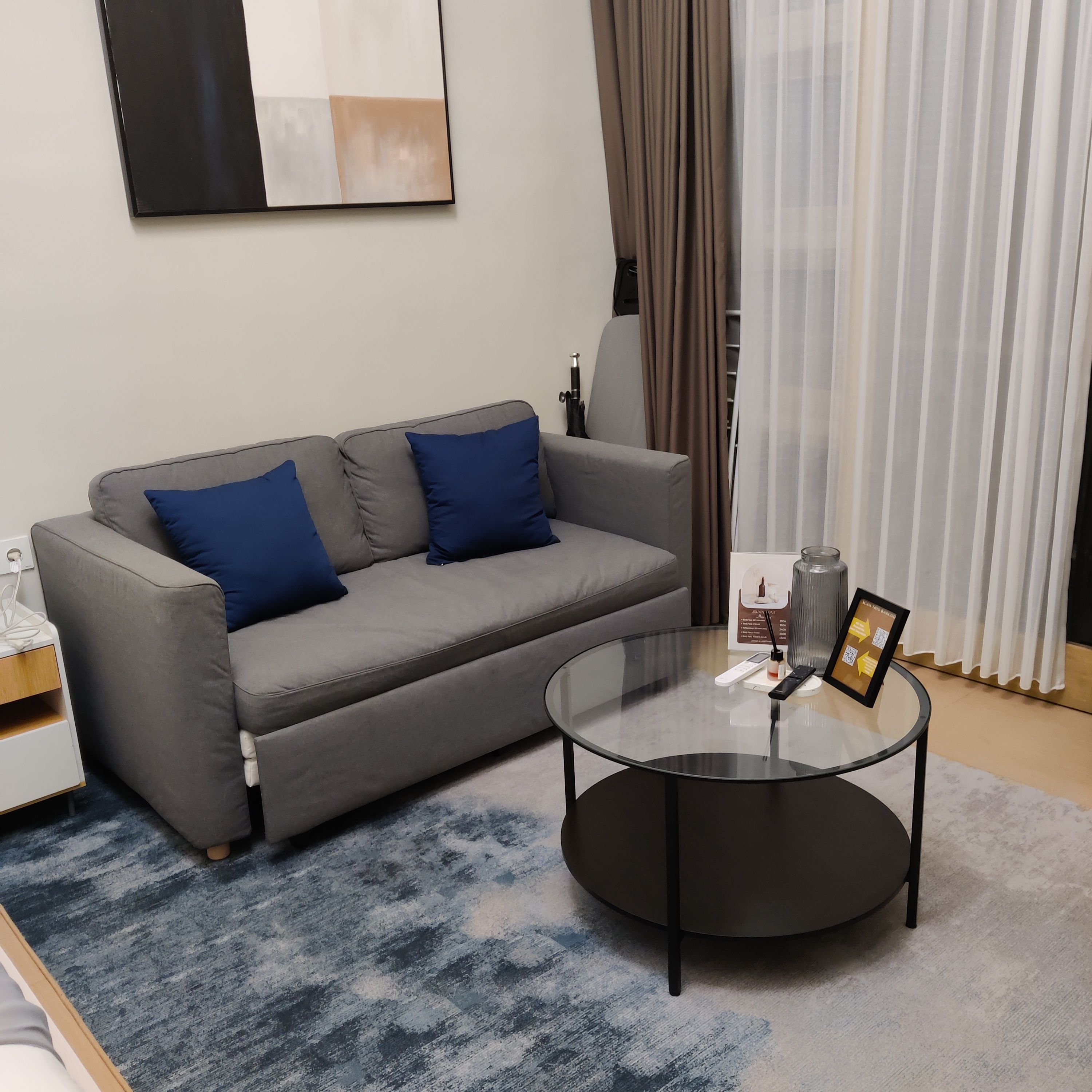 Uma sala de estar moderna com um sofá cinzento, duas almofadas azuis, uma mesa de centro de vidro com um controle remoto, um celular e um quadro emoldurado.
