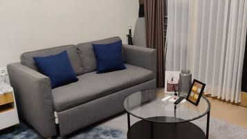 Uma sala de estar moderna com um sofá cinzento, duas almofadas azuis, uma mesa de centro de vidro com um controle remoto, um celular e um quadro emoldurado.