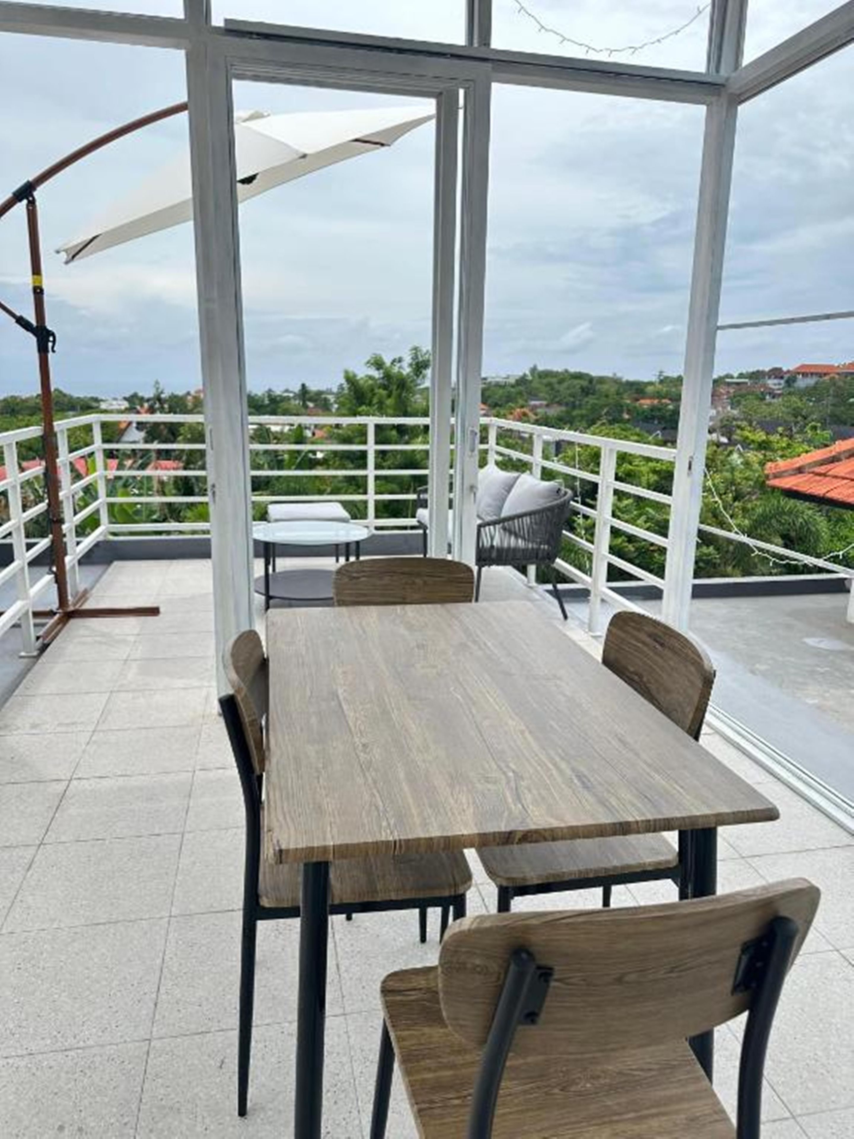 Um pátio externo com mesa e cadeiras de madeira, um guarda-sol branco e vista para o verde ao redor.