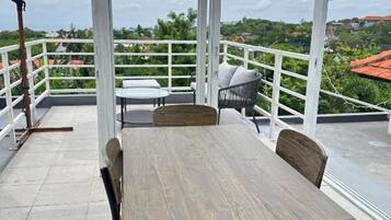 Um pátio externo com mesa e cadeiras de madeira, um guarda-sol branco e vista para o verde ao redor.