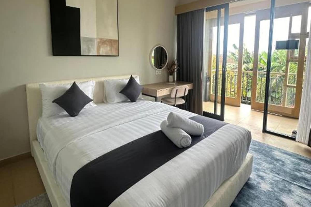 Quarto de hotel com cama grande, escrivaninha, cadeira, espelho redondo e vista para o exterior através de uma porta de correr de vidro.