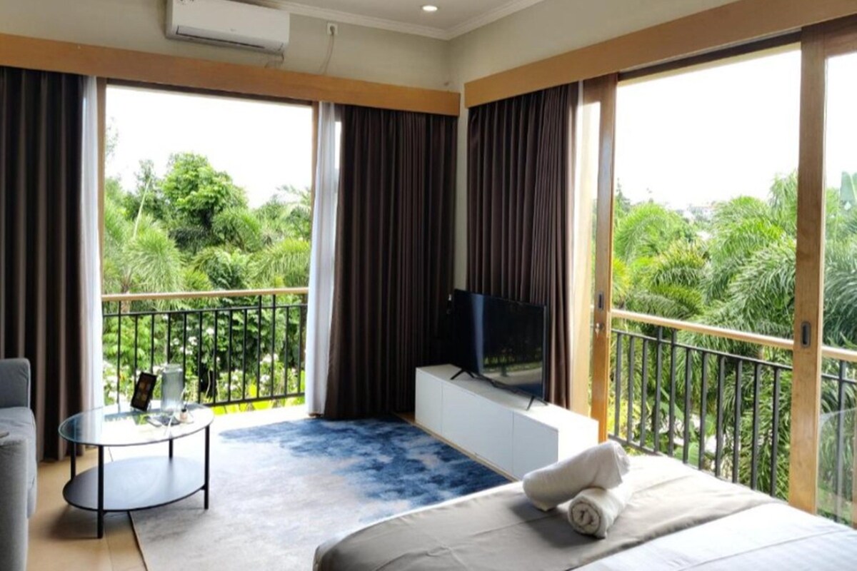 Quarto de hotel com varanda, televisão de tela plana, cama, mesa de centro redonda e vista para área verde.