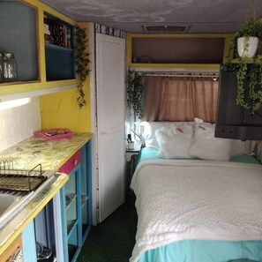 Quarto compacto com cama, escrivaninha pequena, geladeira e janela com cortinas.