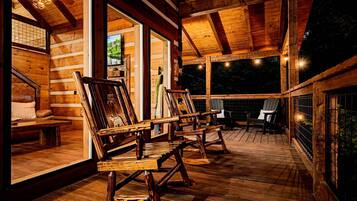 Interior de cabana de madeira com varanda, cadeiras rústicas de madeira e lareira de pedra.