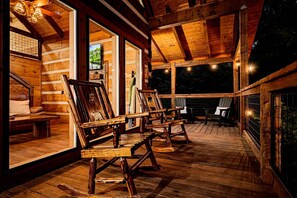 Interior de cabana de madeira com varanda, cadeiras rústicas de madeira e lareira de pedra.