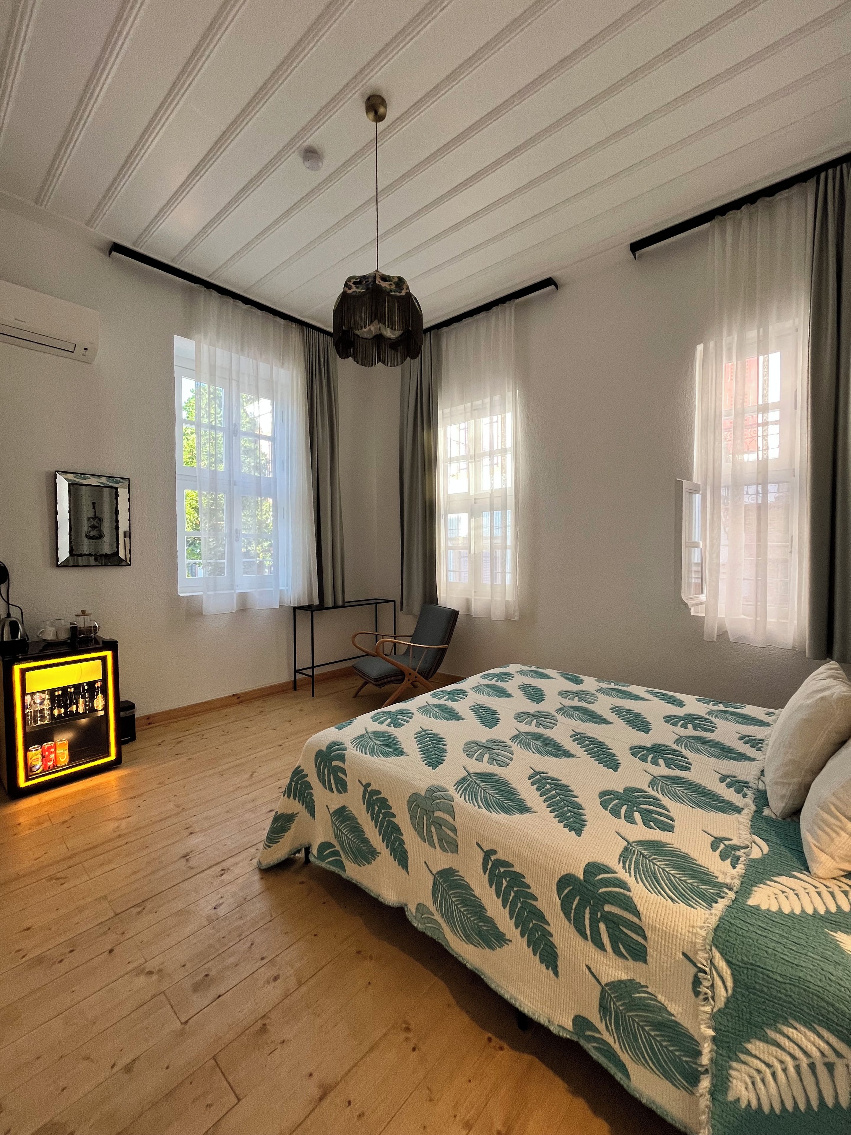 Quarto com cama, cadeira, mesinha de cabeceira com luminária e janela com cortinas.