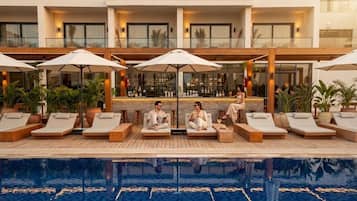 Área da piscina com espreguiçadeiras e guarda-sóis, um bar e reflexos na água.