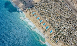 Vista aérea de uma área residencial costeira com praia, piscinas e um conjunto denso de casas.