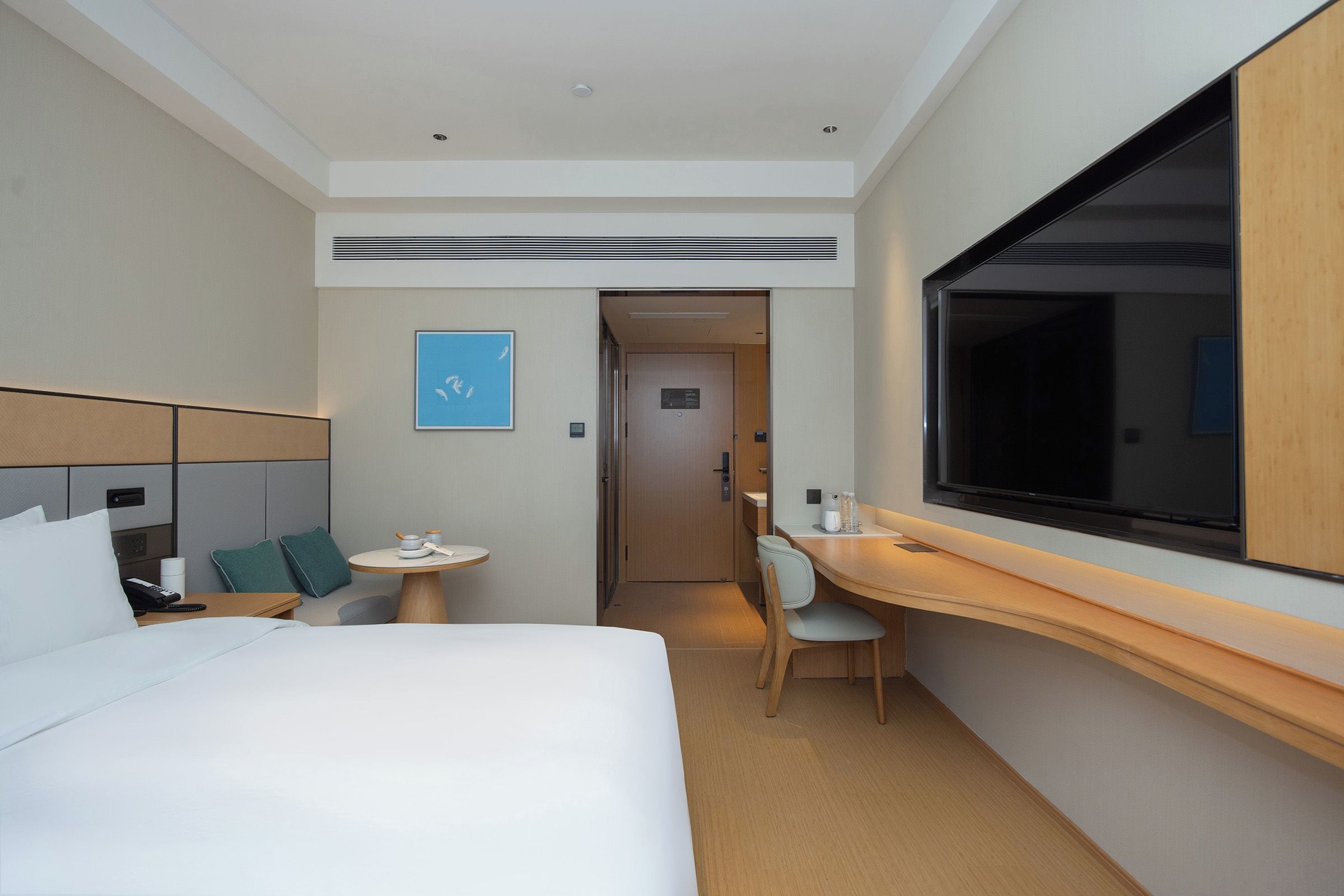 Quarto de hotel com cama, escrivaninha, cadeira, televisão e uma pequena mesa com xícara e telefone.