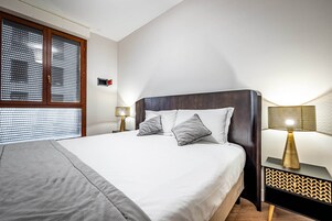 Cama bem arrumada com manta cinza, duas almofadas e cabeceira de madeira. Há uma mesa de cabeceira com abajur e um despertador fixado na parede.
