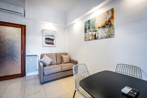Uma sala de estar moderna com um sofá bege, uma mesa de jantar preta e uma cadeira de metal. Há um quadro na parede e um radiador embaixo.