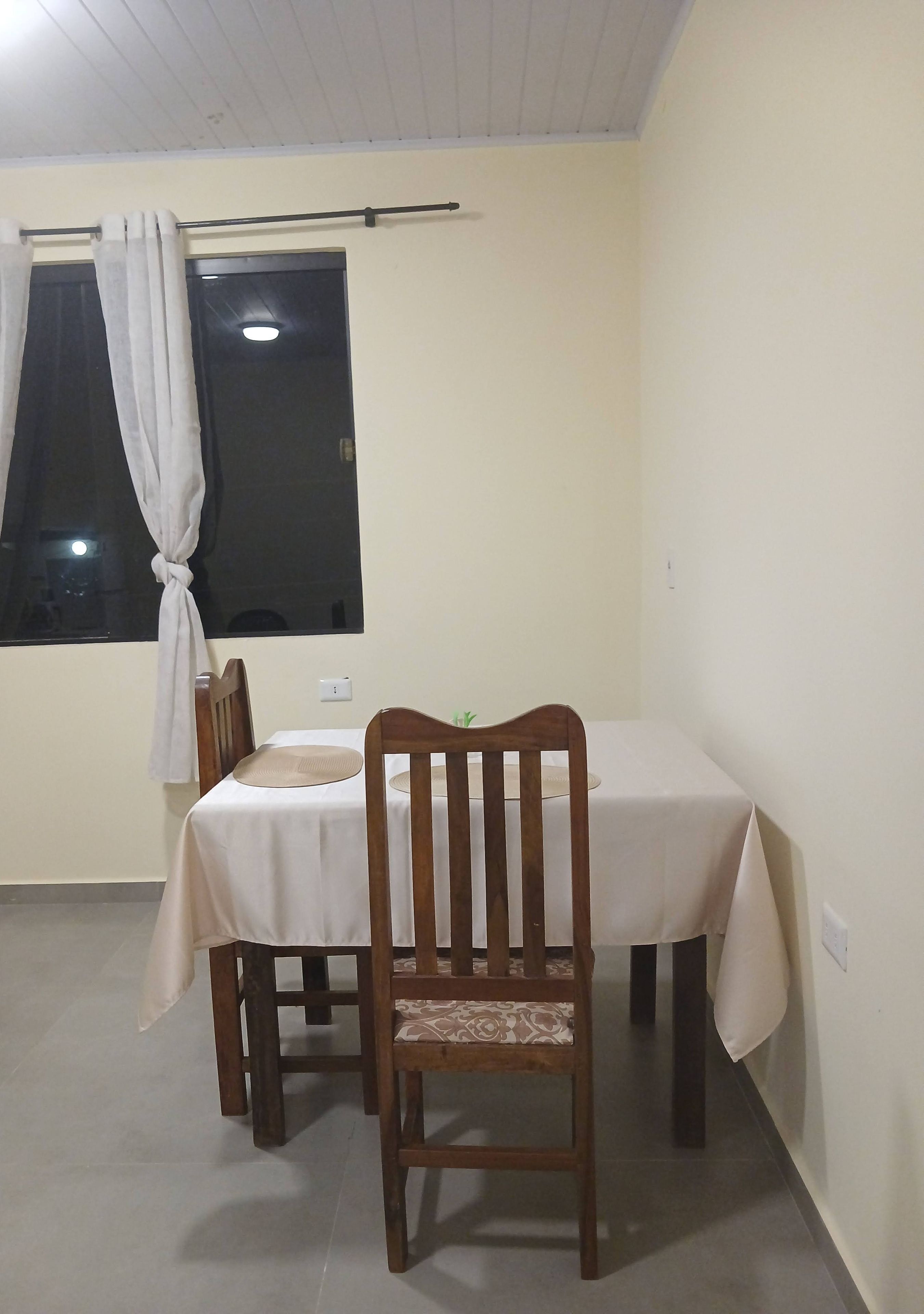Uma área de refeições com mesa e cadeiras, uma janela com cortinas e uma luminária no teto.