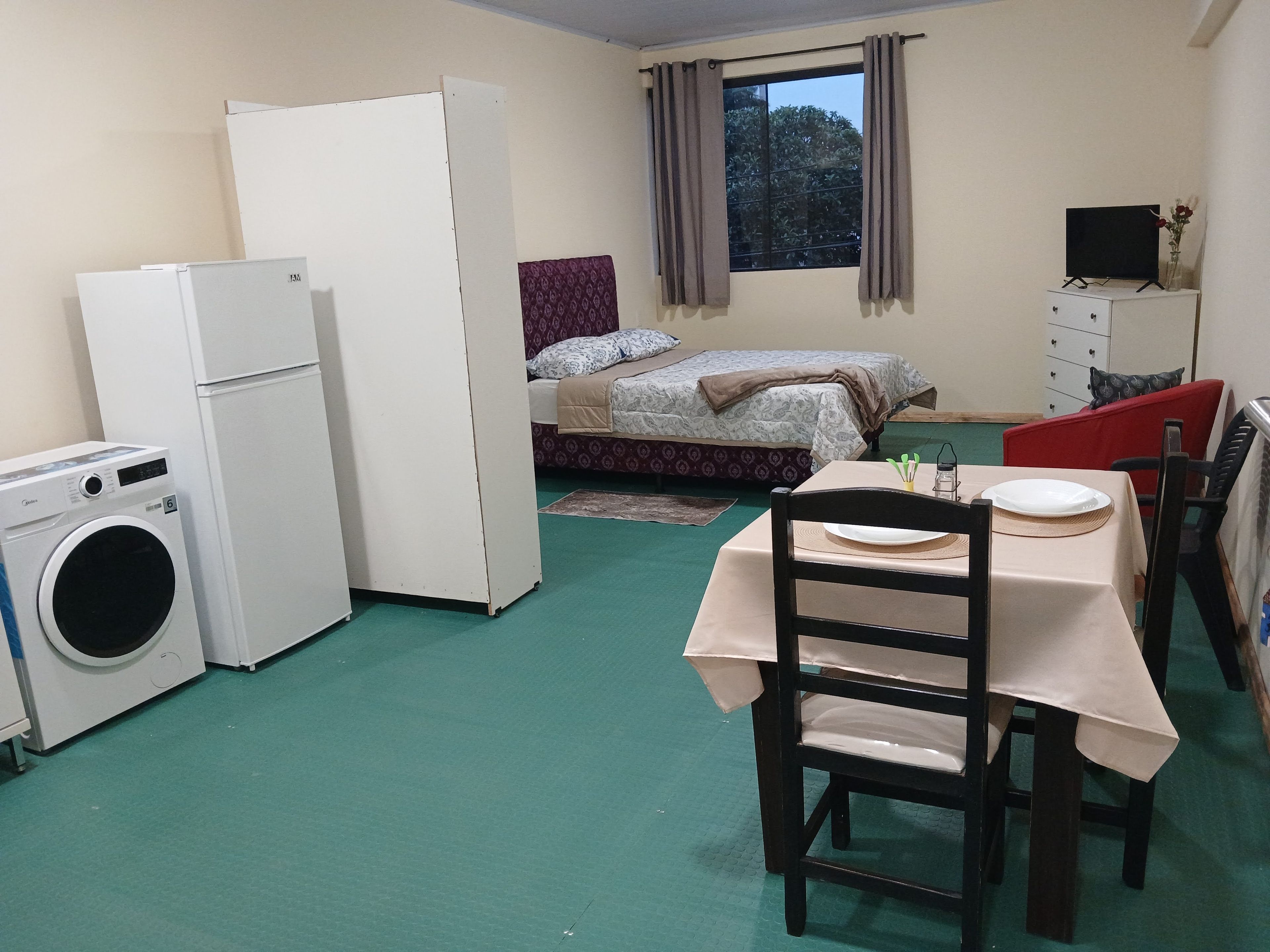 Quarto compacto com cama, mesa, cadeiras, máquina de lavar e televisão.