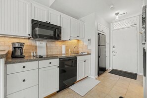 Uma cozinha com armários brancos, micro-ondas preto, máquina de café, lava-louças e frigorífico.