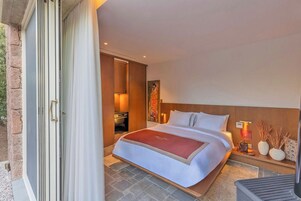Quarto de hotel com uma cama grande, porta de correr para uma varanda, mesa pequena com luminária e um quadro na parede.