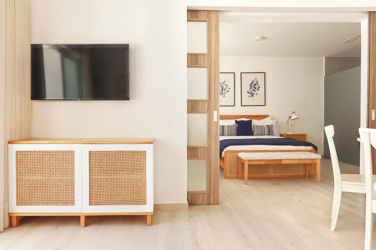 Quarto moderno com televisão de tela plana, mesa de cabeceira de madeira, cama com colcha azul e branca, e uma cadeira branca.