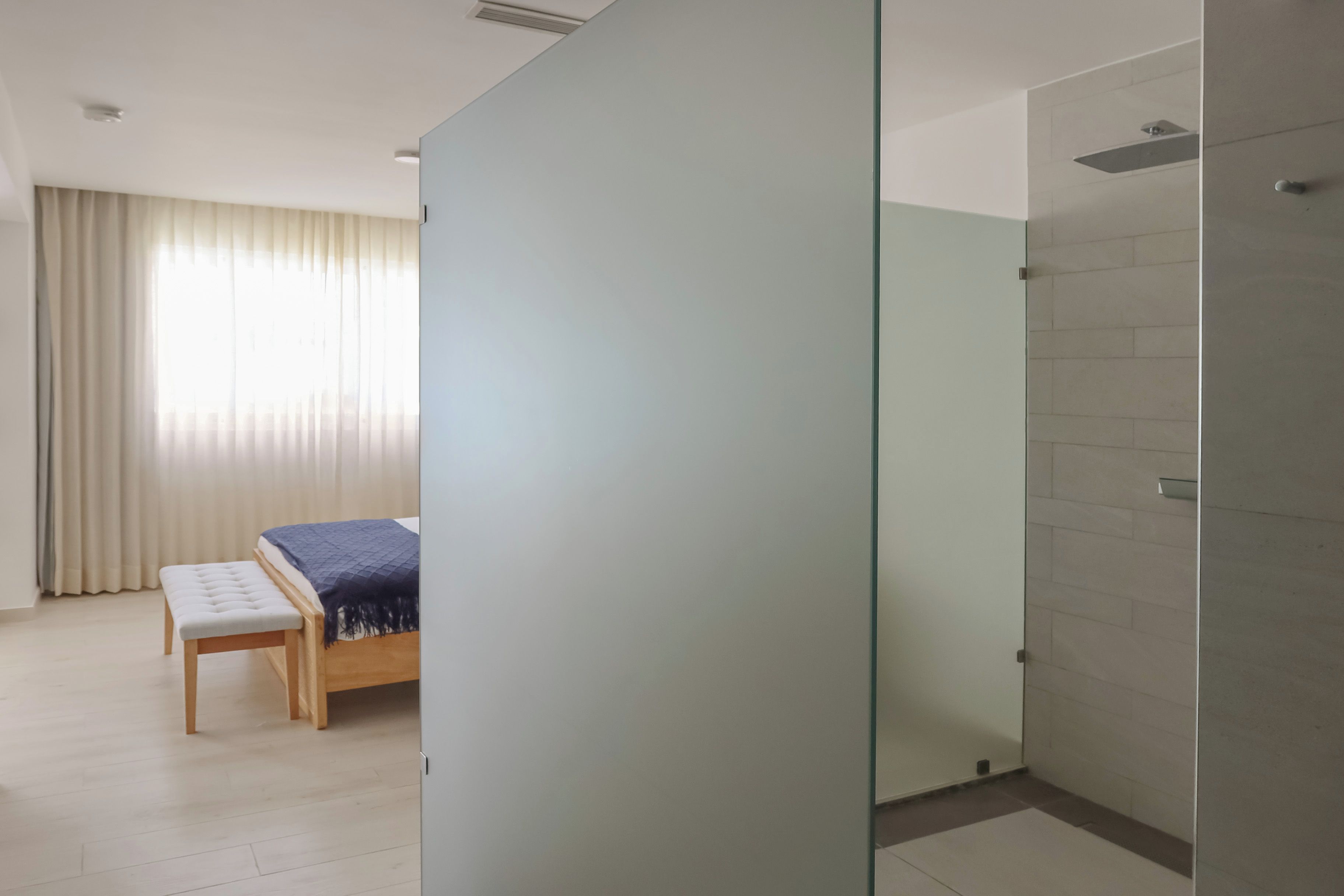 Quarto moderno com cama, um banquinho, divisória de vidro e área de chuveiro.