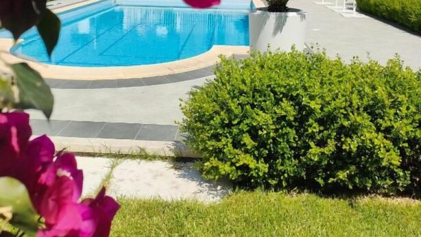Piscina rodeada por vegetação, um edifício com varanda e céu claro.