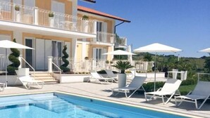 Piscina com espreguiçadeiras e guarda-sóis, um edifício com varandas e um céu claro.