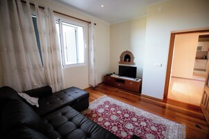 Uma sala de estar com um sofá preto, uma televisão sobre um suporte de madeira, um tapete estampado e uma janela com cortinas.
