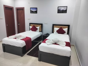 Quarto de hotel com duas camas, cada uma com cabeceira, roupa de cama branca e almofadas vermelhas. Há duas portas, um interruptor de luz fixado na parede e dois quadros na parede.