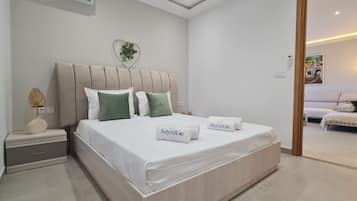 Quarto moderno com uma cama grande, uma mesinha de cabeceira, um detalhe decorativo em formato de coração na parede e vista para outro cômodo.