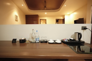 Quarto de hotel com escrivaninha de madeira, cafeteira, chaleira, bandeja com xícaras e garrafa de água, e uma caixa Nestlé.