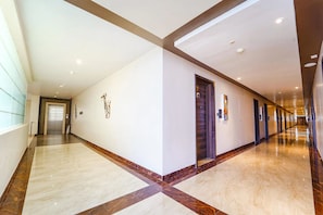 Um corredor longo e bem iluminado, com piso de mármore, portas e alguns objetos fixados na parede.
