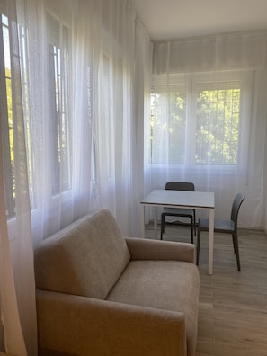 Quarto com sofá bege, mesa branca e duas cadeiras. Janelas amplas com cortinas leves.