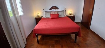 Uma cama com colcha vermelha, duas mesas de cabeceira com abajures e piso de madeira.