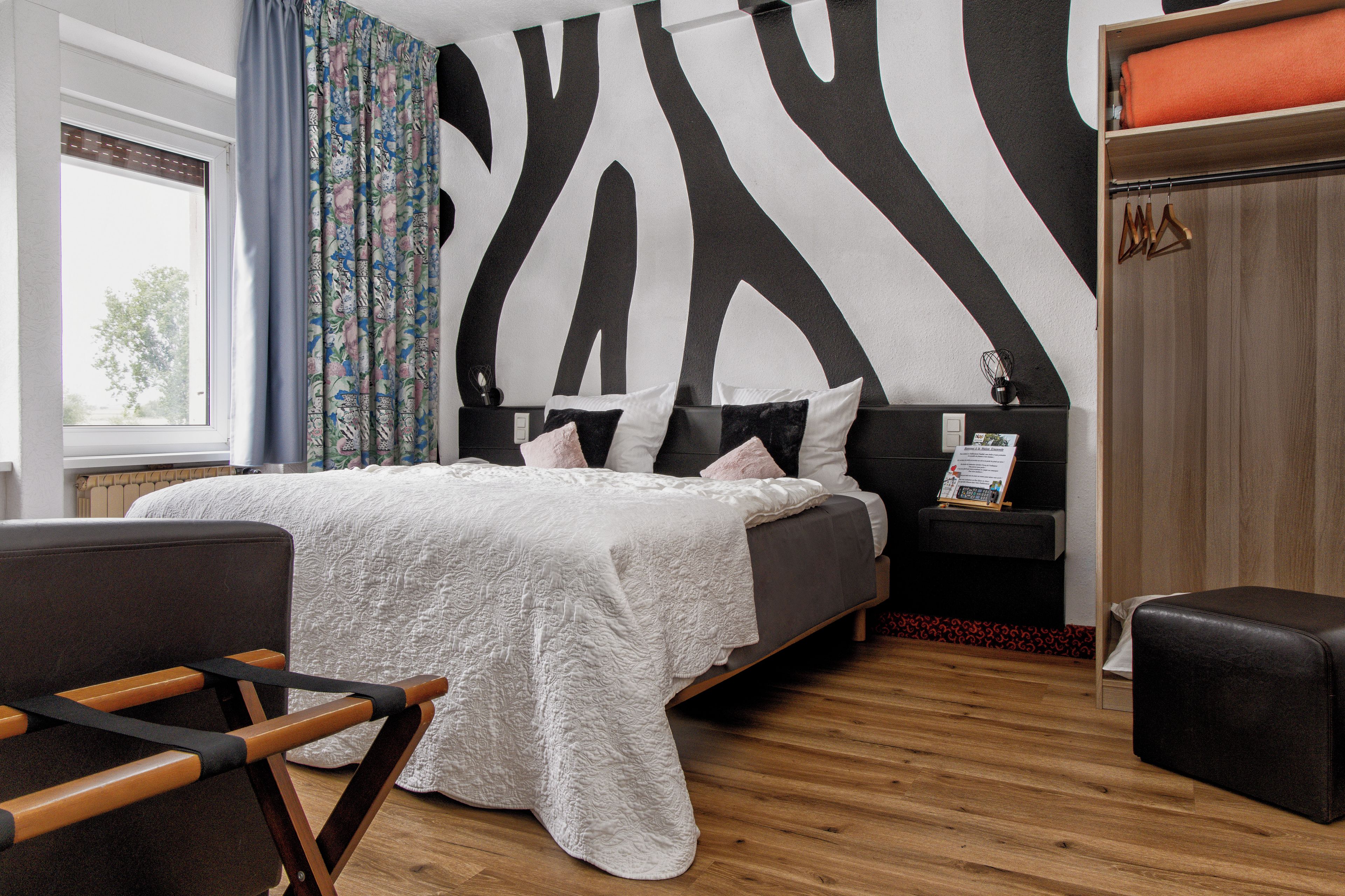 Quarto moderno com cama grande, piso de madeira, parede com padrão de zebra e janela com cortinas.