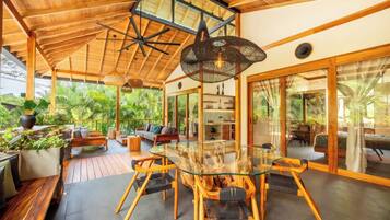 Área externa ampla com mesa de vidro rodeada por cadeiras, teto de madeira e vista para vegetação exuberante.