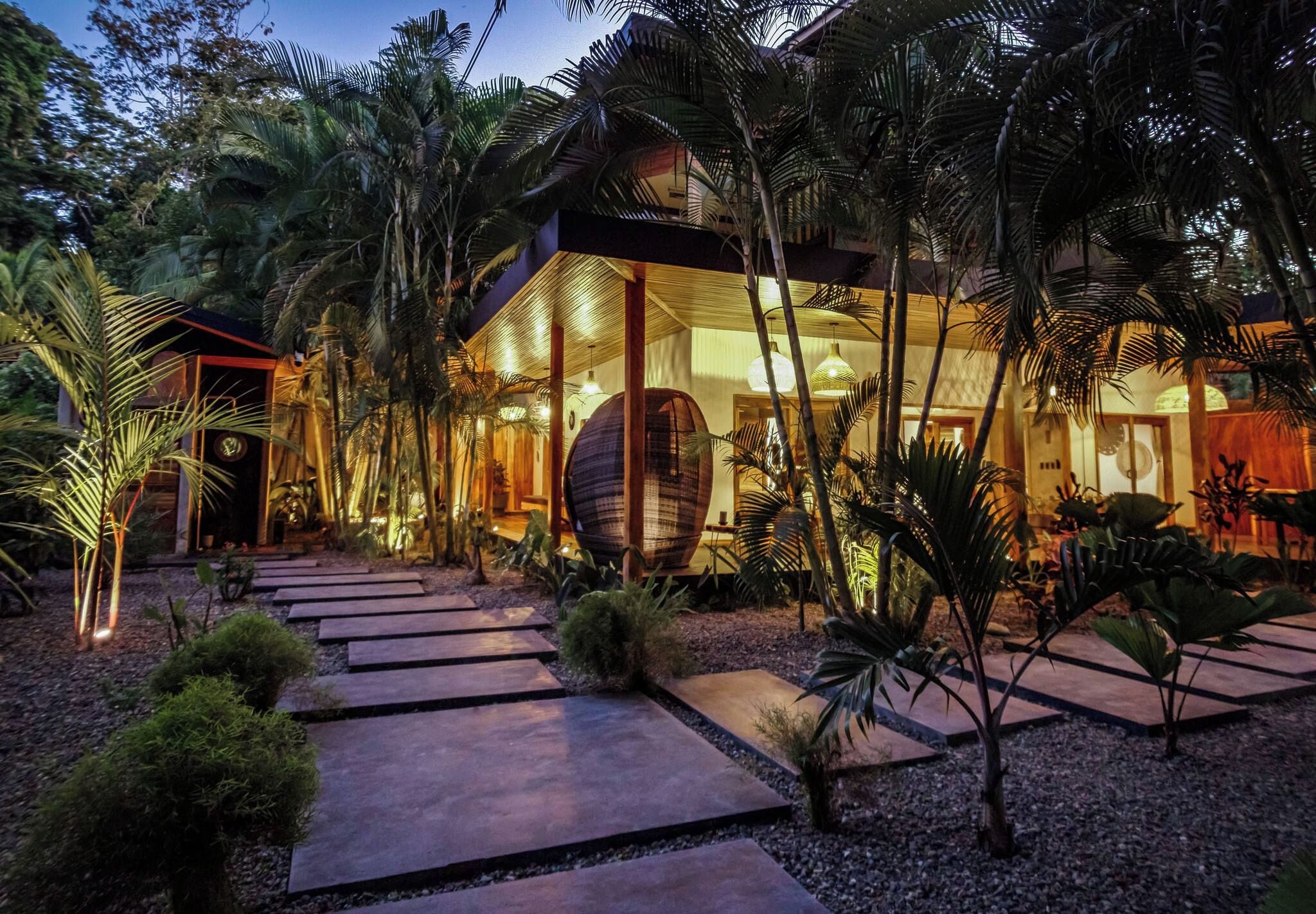 Uma casa moderna com fachada de vidro, rodeada por plantas tropicais e um caminho de pedra.