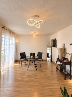 Um quarto amplo com uma mesa redonda, quatro cadeiras, um lustre no teto, um abajur e um armário de madeira.