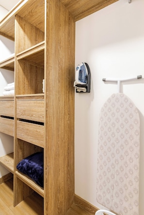 Um guarda-roupa de madeira com várias prateleiras e espaço para pendurar, uma tábua de passar branca com capa estampada, e uma pequena caixa com ferro ao lado na parede.