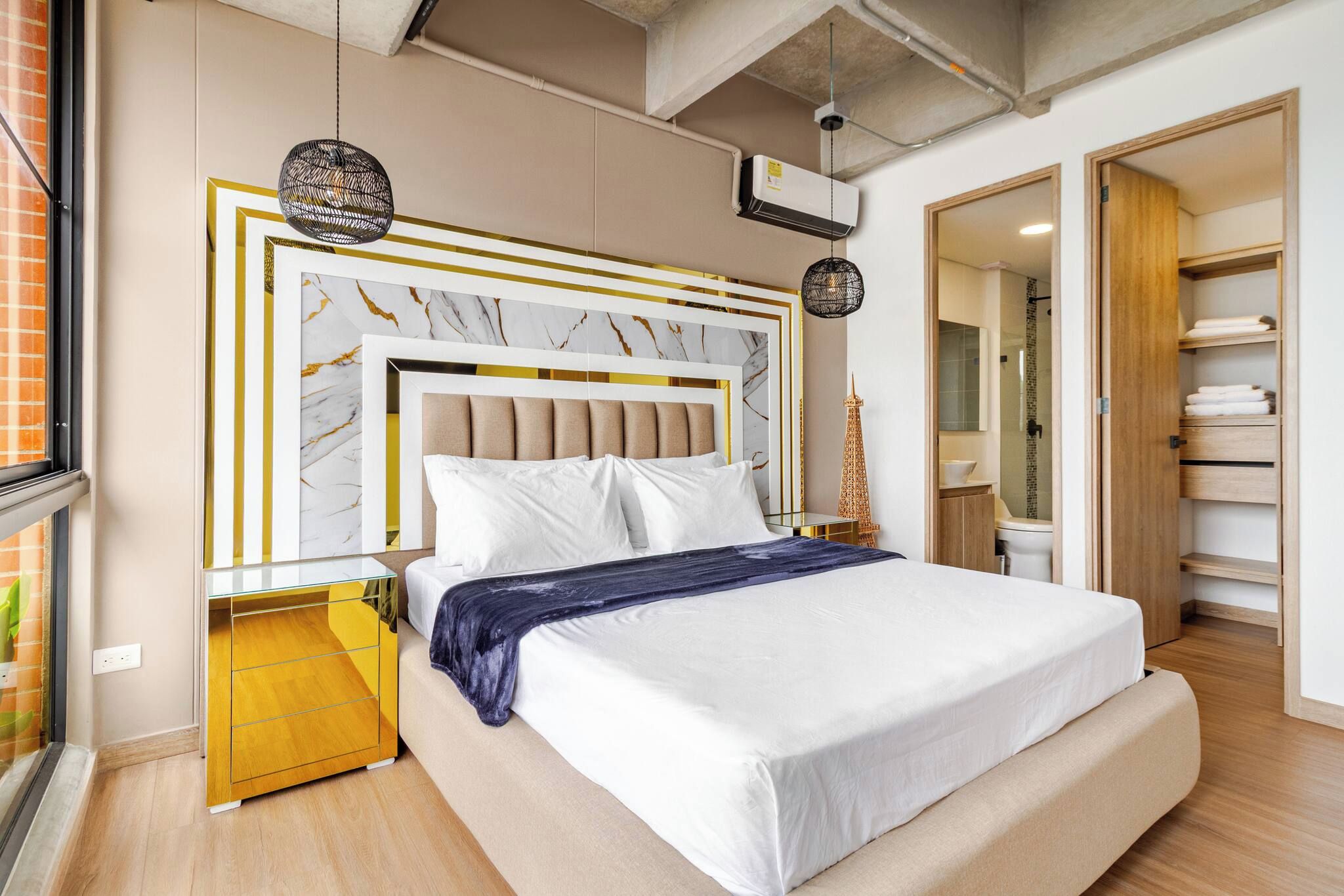 Quarto moderno com cama grande, mesa de cabeceira, guarda-roupa e banheiro visível por uma porta aberta.