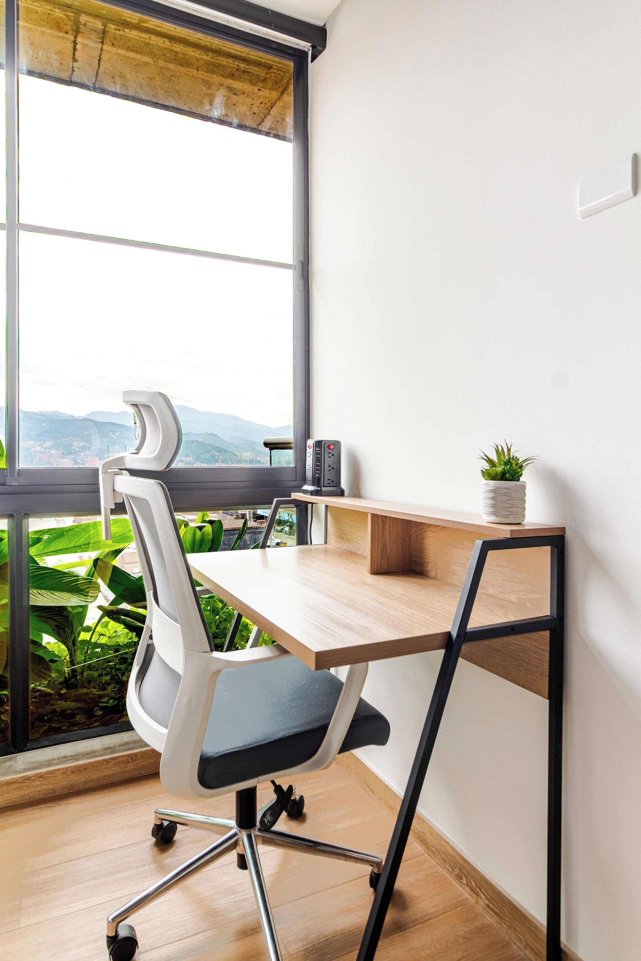Um espaço de escritório moderno com escrivaninha, cadeira e vista para o verde através de uma grande janela.