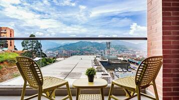 Varanda com duas cadeiras de vime e uma mesa pequena, com vista para paisagem urbana com edifícios e montanhas.