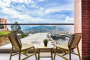 Varanda com duas cadeiras de vime e uma mesa pequena, com vista para paisagem urbana com edifícios e montanhas.