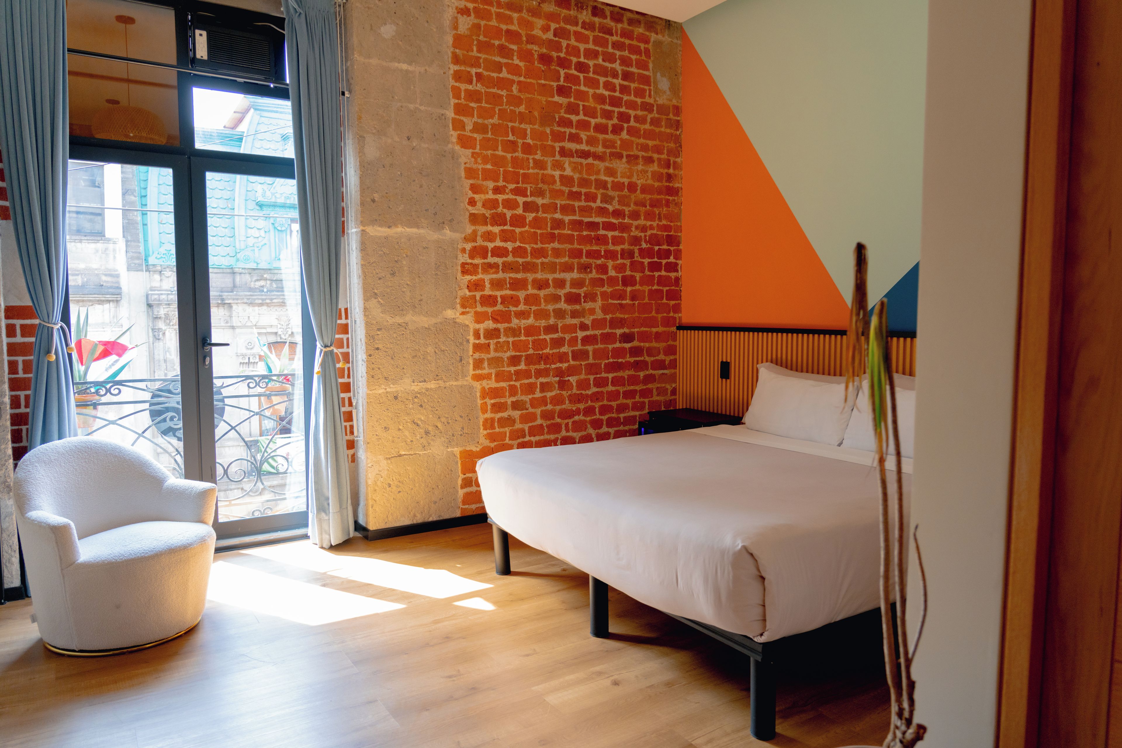 Quarto de hotel com cama, uma cadeira, janela com cortinas e parede de tijolos.